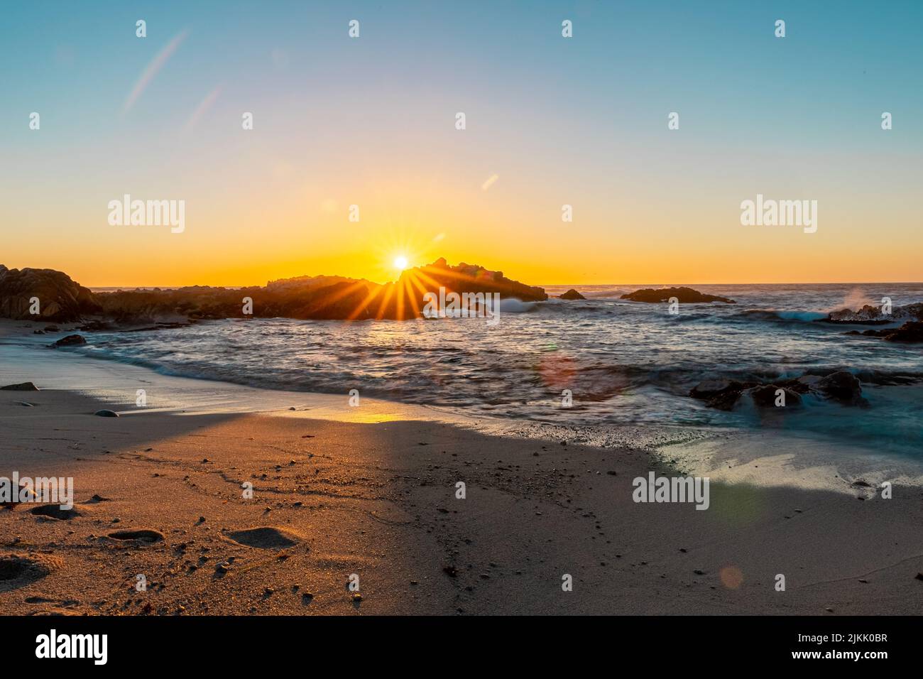 A scenic view of sun from behind the rocks over the water and the sand ...