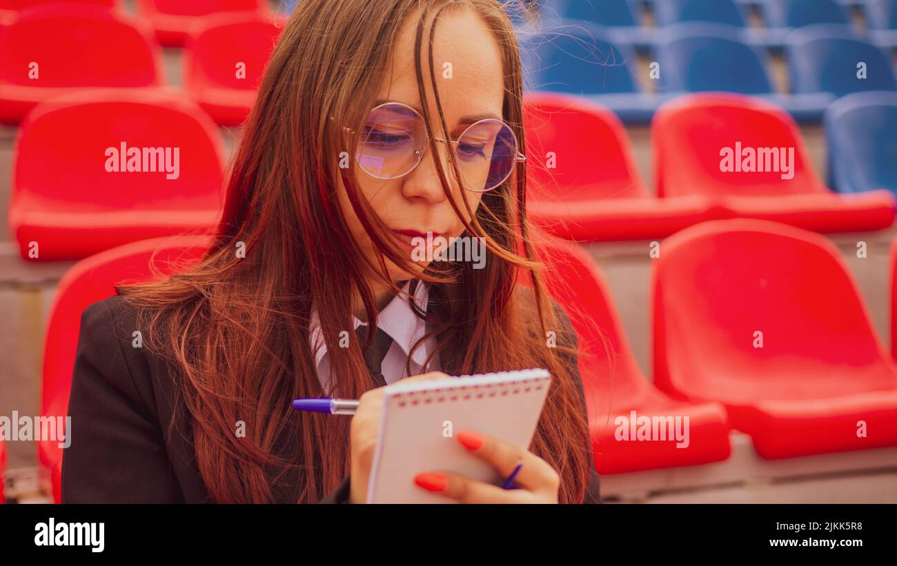 Young woman in glasses with notepad, pen sitting on stadium bleachers ...