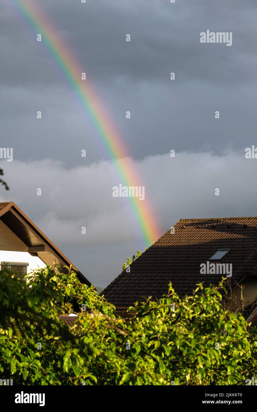 A vertical shot of a rainbow going over roofs of houses and a cloudy ...