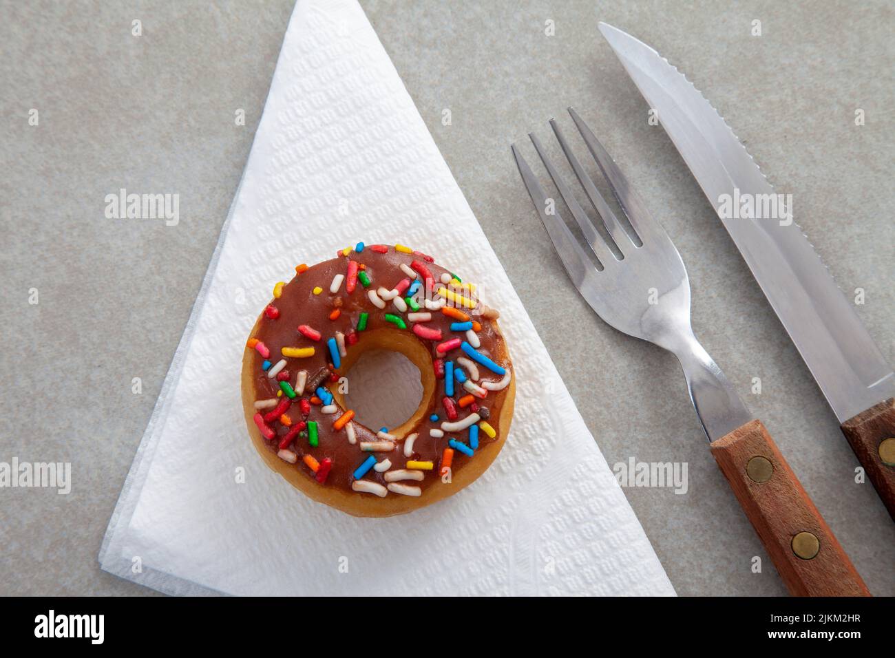 A top view of a chocolate sprinkle donut on a tissue and silverware ...