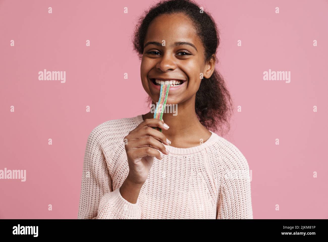 Brunette black girl smiling while eating candy worm isolated over pink ...