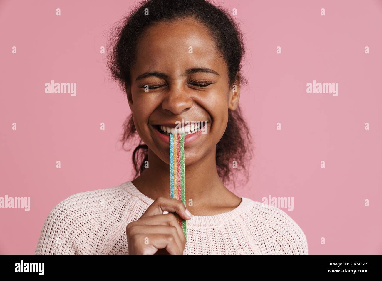 Brunette black girl smiling while eating candy worm isolated over pink ...