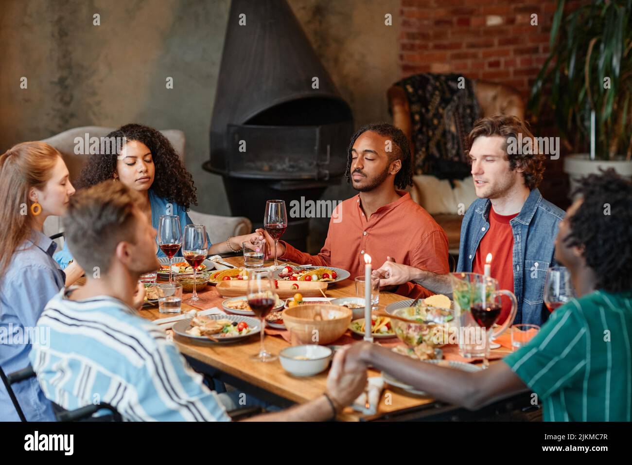 Diverse group of young people holding hands at table and saying grace ...