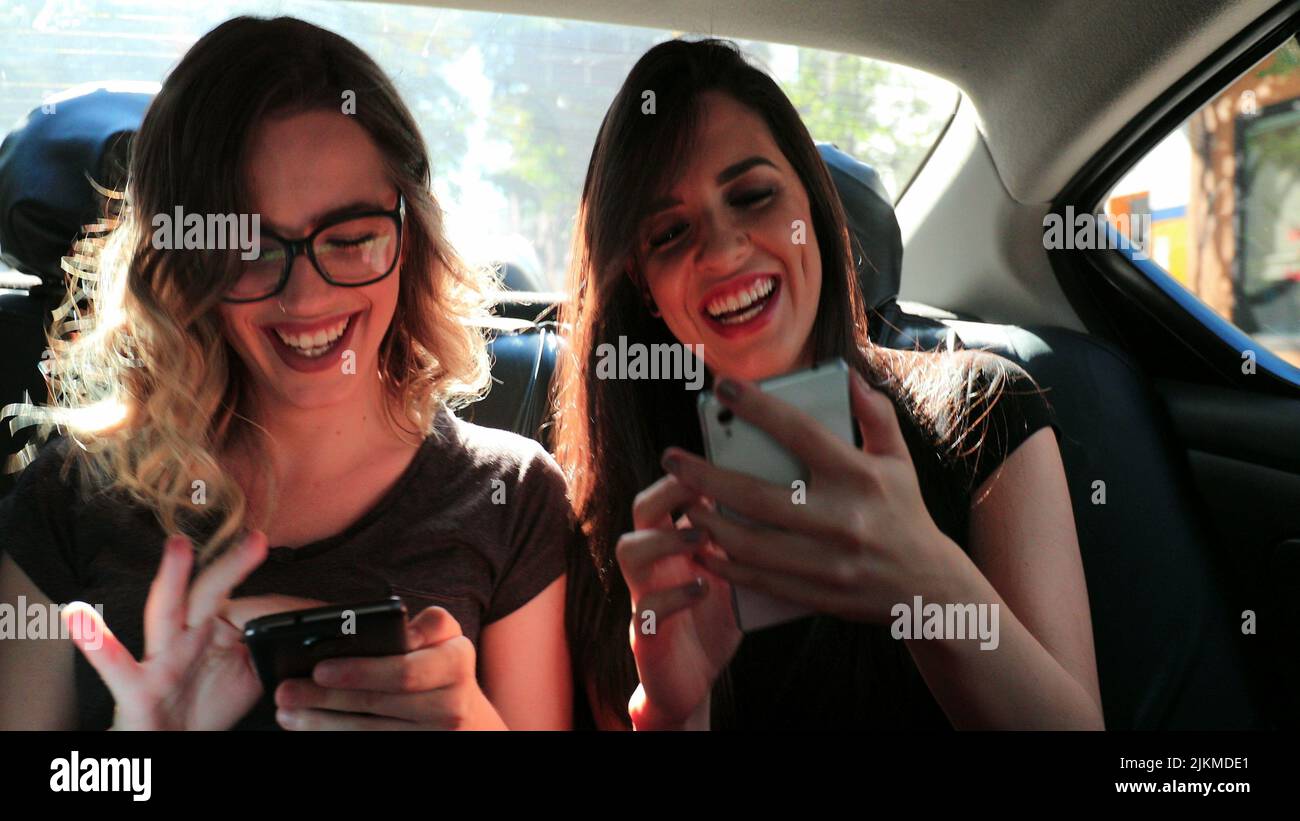 Female friends browsing on their smartphone while riding cab laughing ...