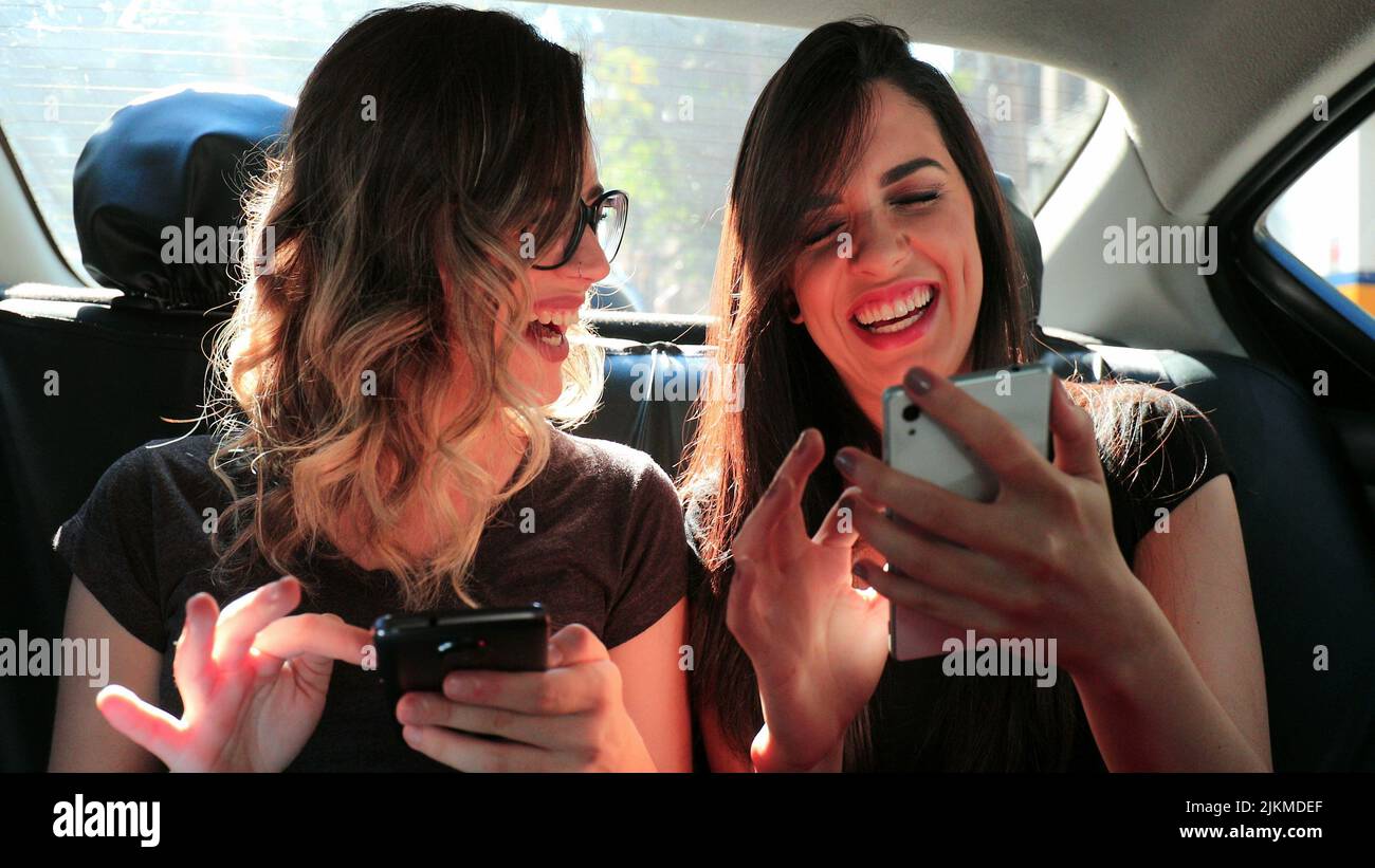 Female friends browsing on their smartphone while riding cab laughing ...