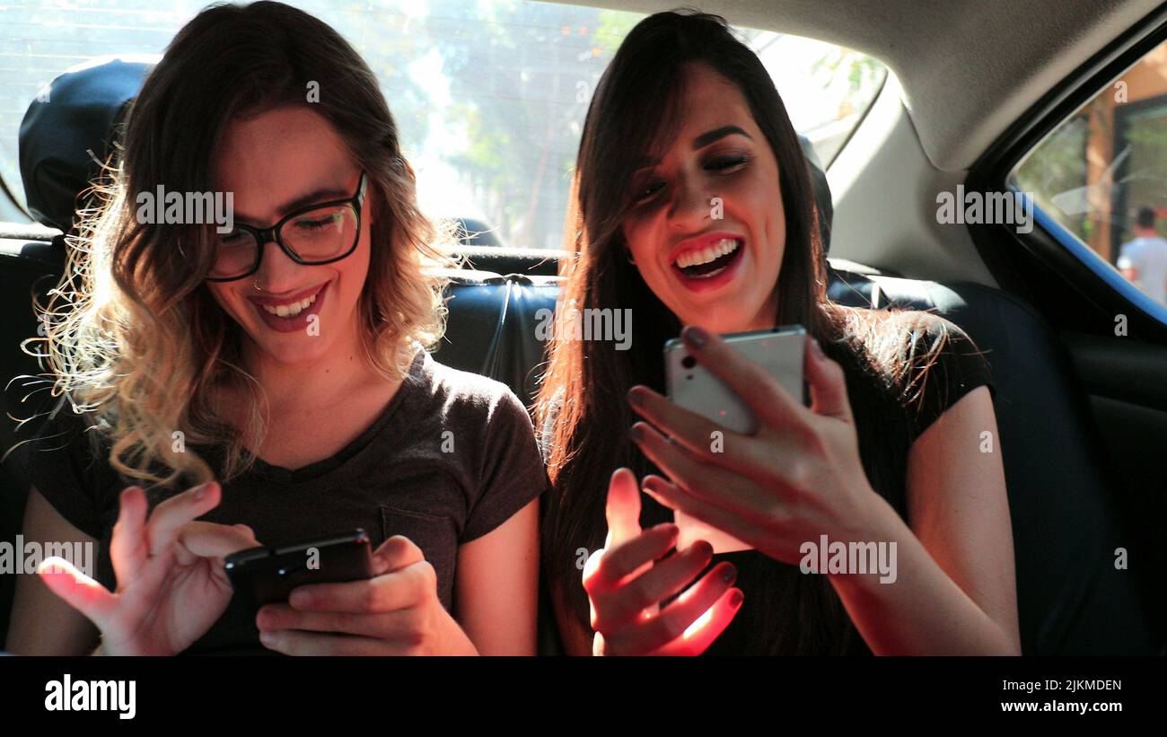 Female Friends Browsing On Their Smartphone While Riding Cab Laughing