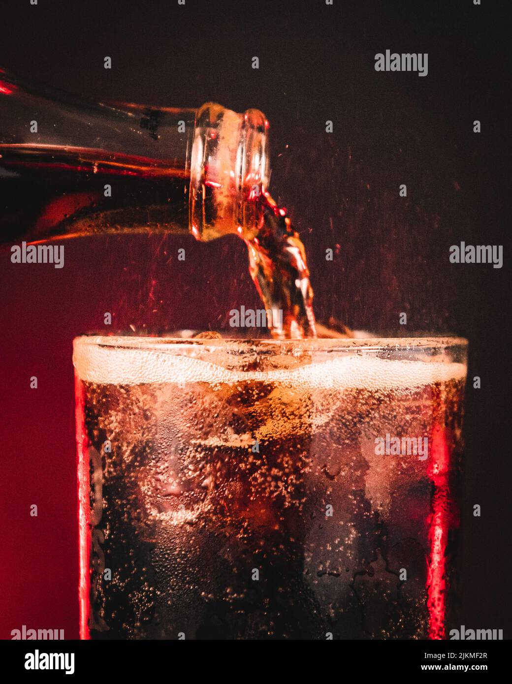 A vertical closeup of a bottle pouring soda into a with ice cubes in it ...