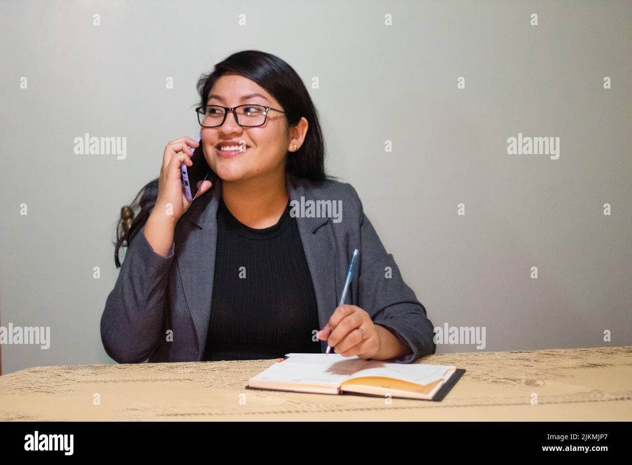 A caucasian female in a suit taking with her phone and writing notes ...