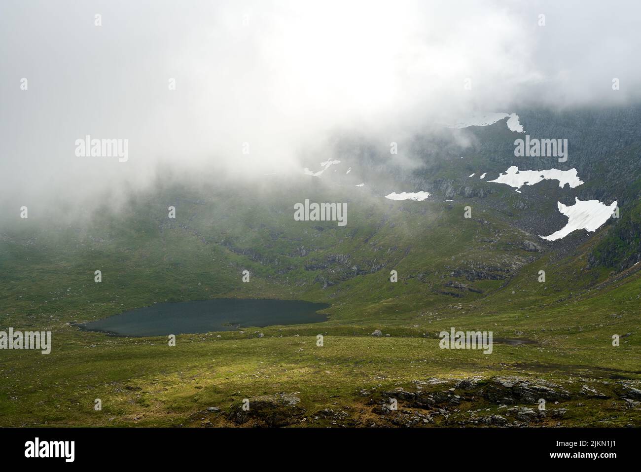 A scenic view of a small lake, snow spots, and foggy mountains during ...
