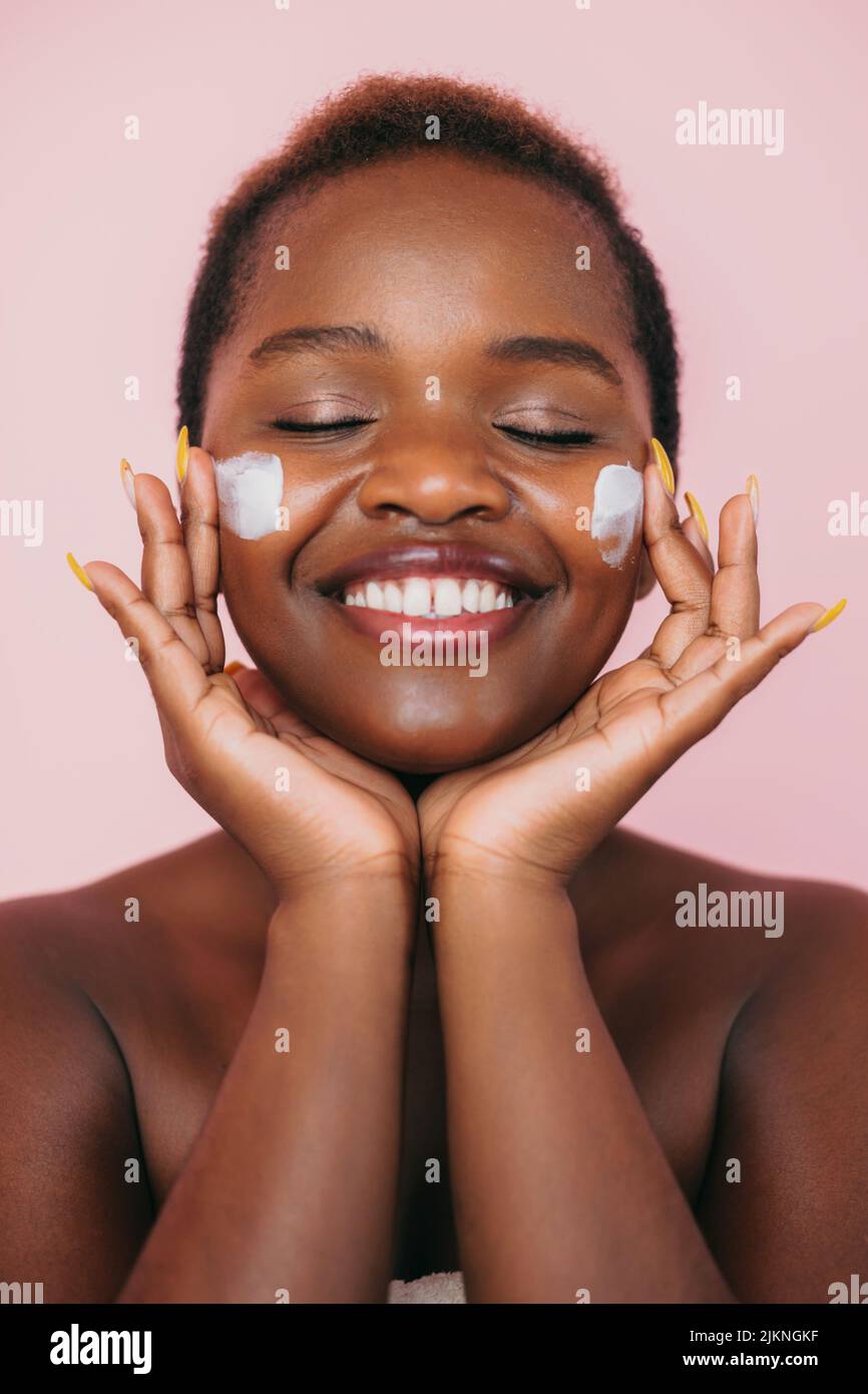 Portrait of smiling black woman keeping her eyes closed applying face ...