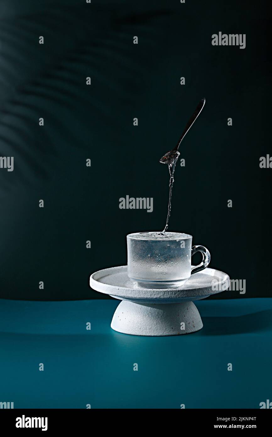 A cup with cold liquid with levitating spoot on a cyan table near black ...