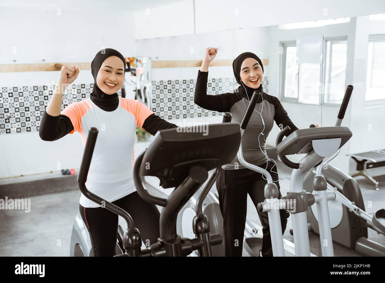 excited muslim woman exercising with friend on static elliptical cycle ...