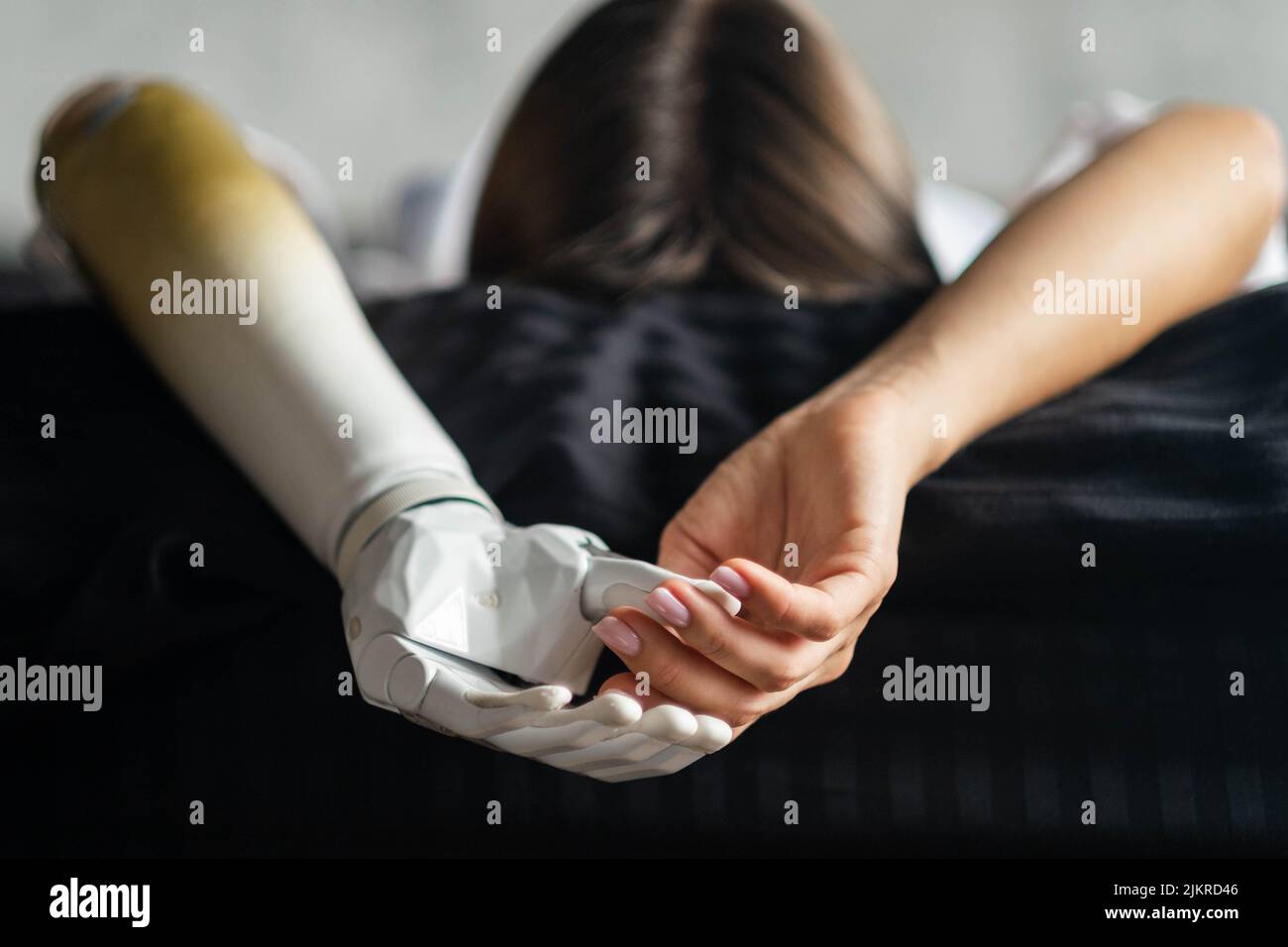 Close up two hands of woman with bionic arm on black bed at home ...