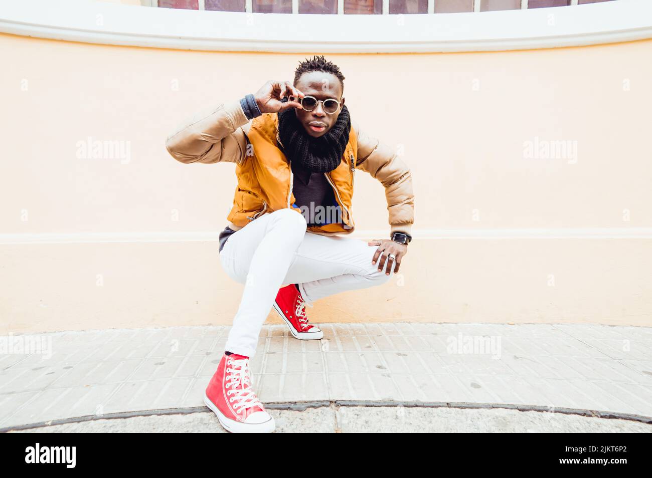 stylish african man in the street crouching with white wall behind him ...