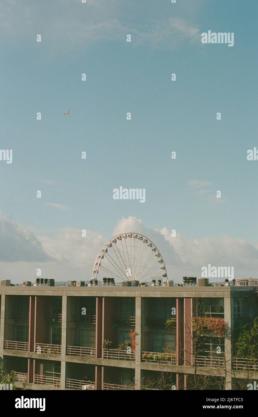 A vertical view of a Ferris wheel behind a building on a blue cloudy ...