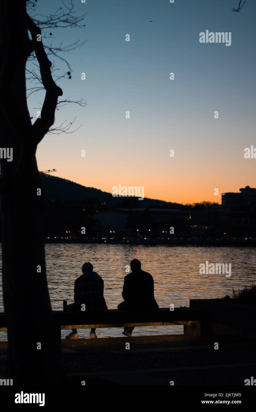 The silhouette of two people sitting on the bench on the coast at ...