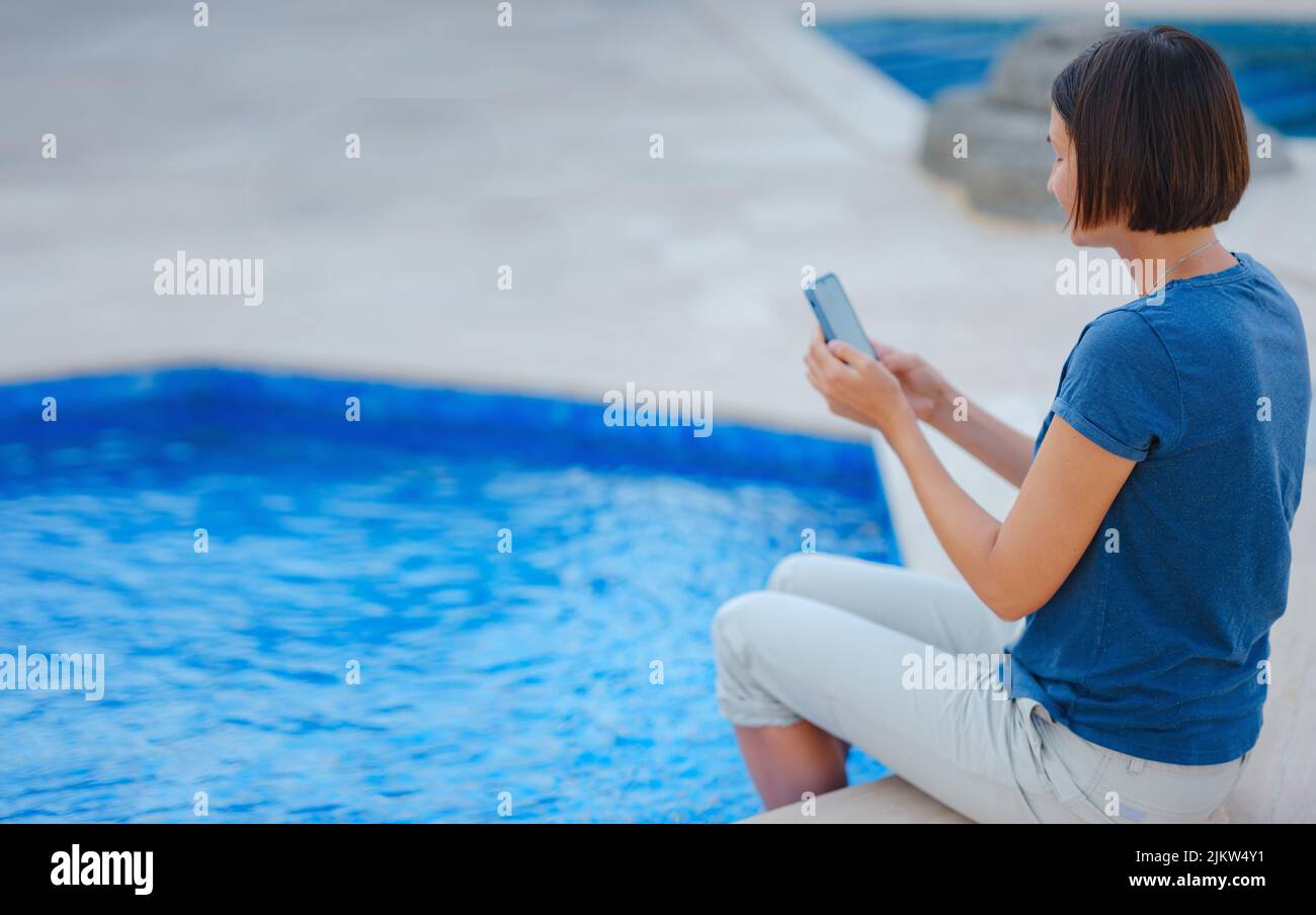 Young brunette woman using her phone while relaxing by the pool . Young ...
