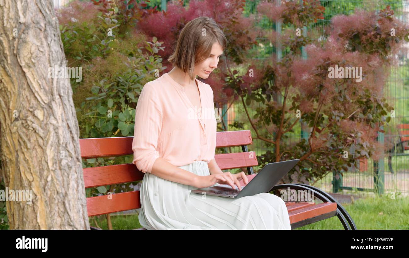 Caucasian young woman sitting on a bench in a park and working on her ...