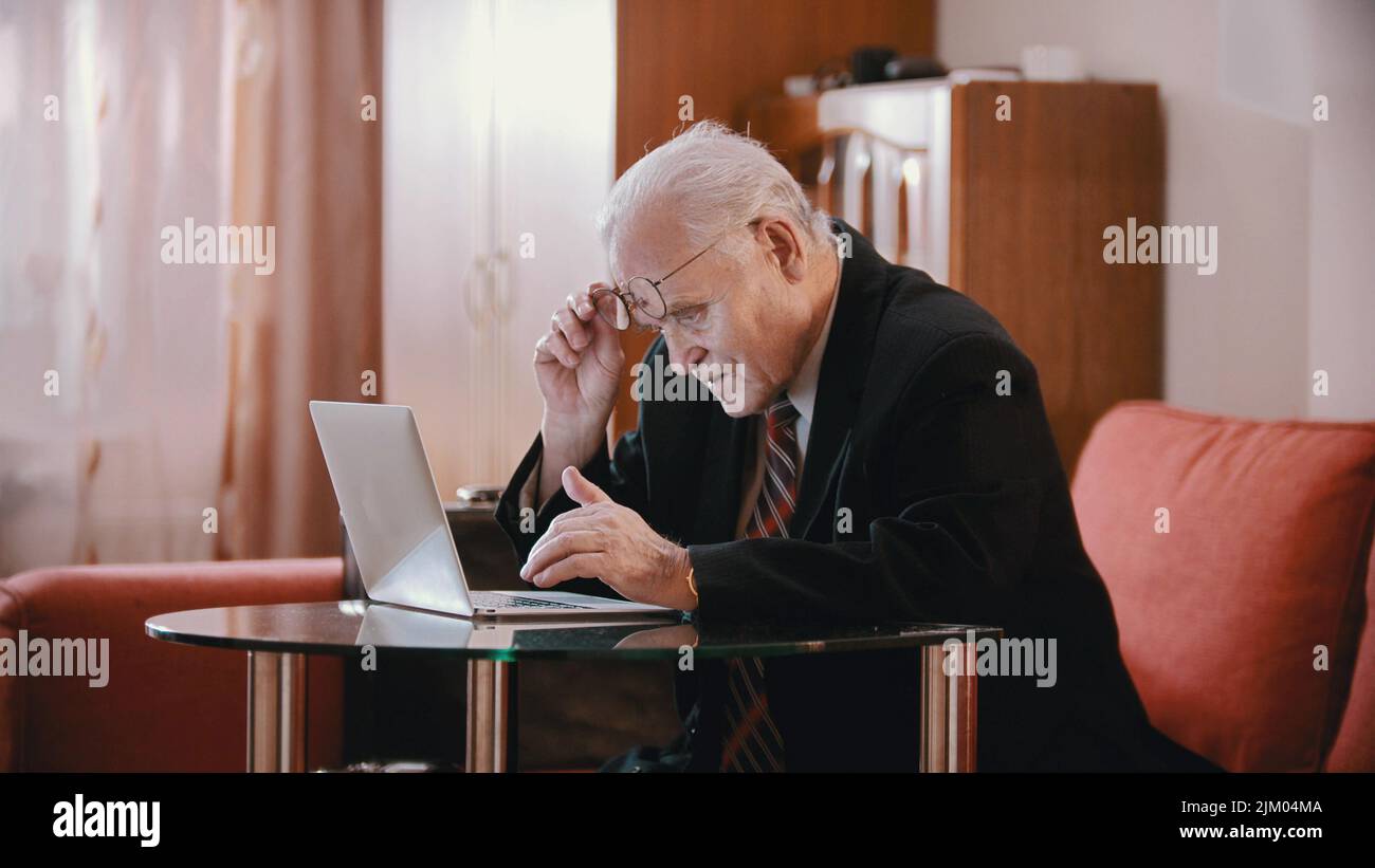 An old man in glasses sitting by the laptop at looking at the monitor ...
