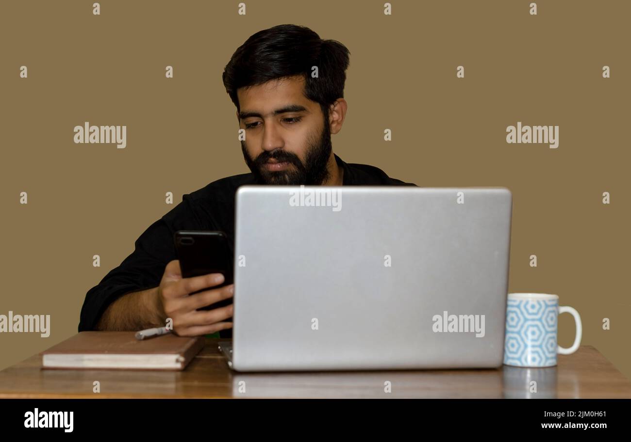 A young indian man looking at phone while working on laptop. Selective ...