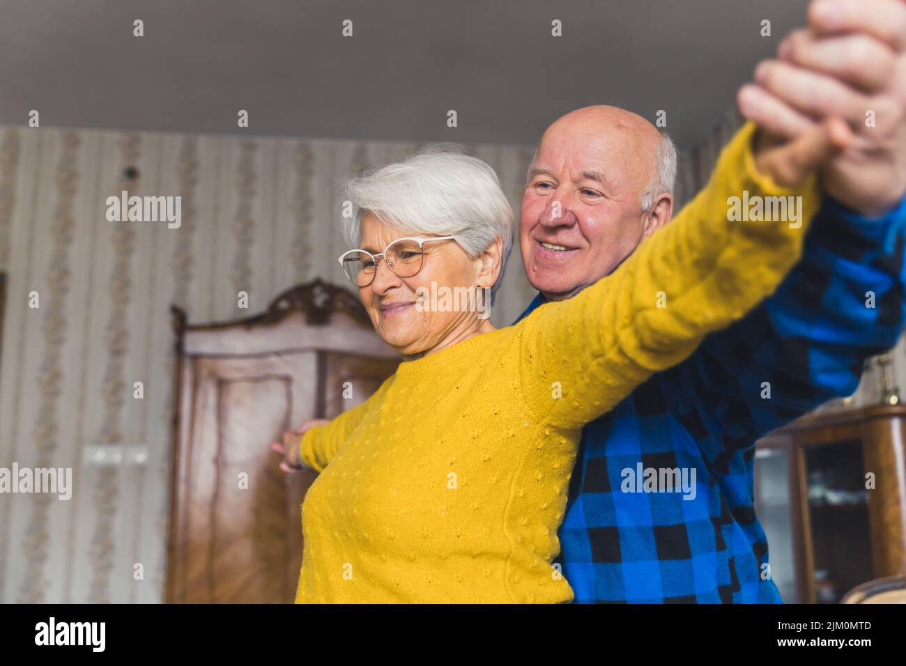 Cute joyful senior European married couple dancing, smiling, and having fun in the living room ...