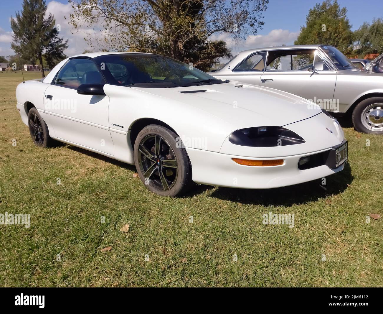 Old white sport Chevrolet Camaro coupe 1990s by GM in the countryside. Nature, grass, trees ...