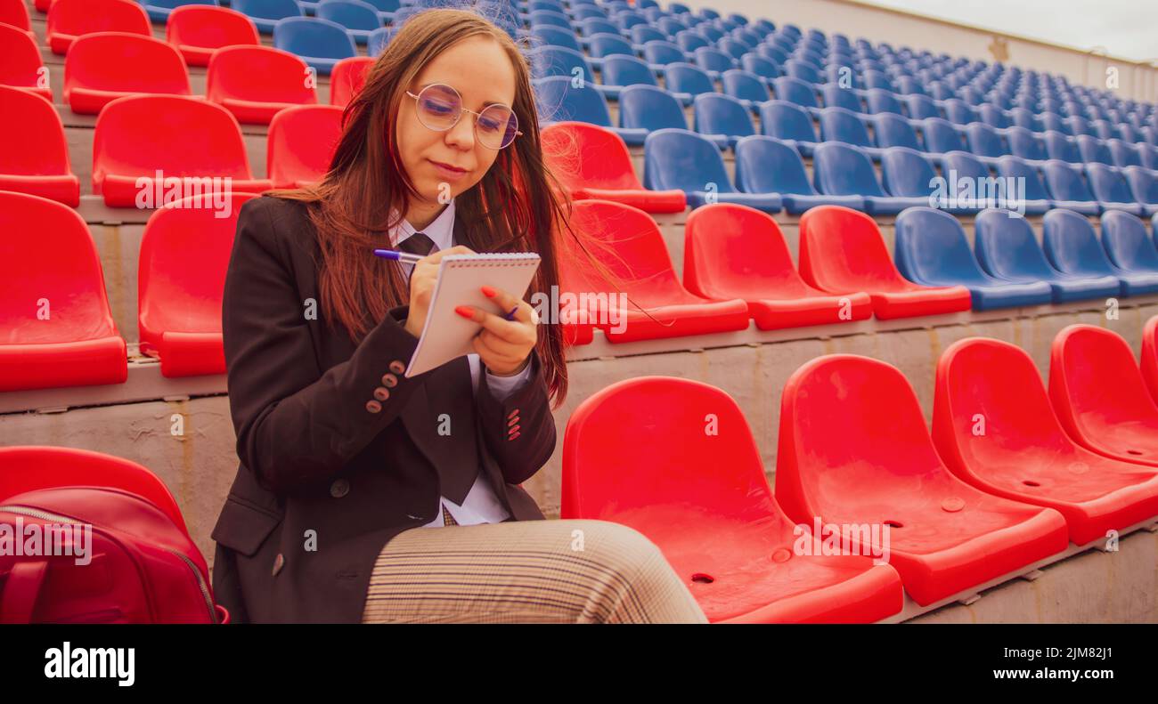 Young woman in glasses with notepad, pen sitting on stadium bleachers ...