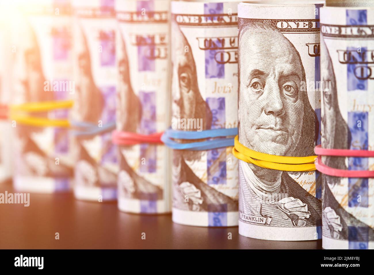 A row of rolled hundred dollar bills with rubber bands with sunlights ...