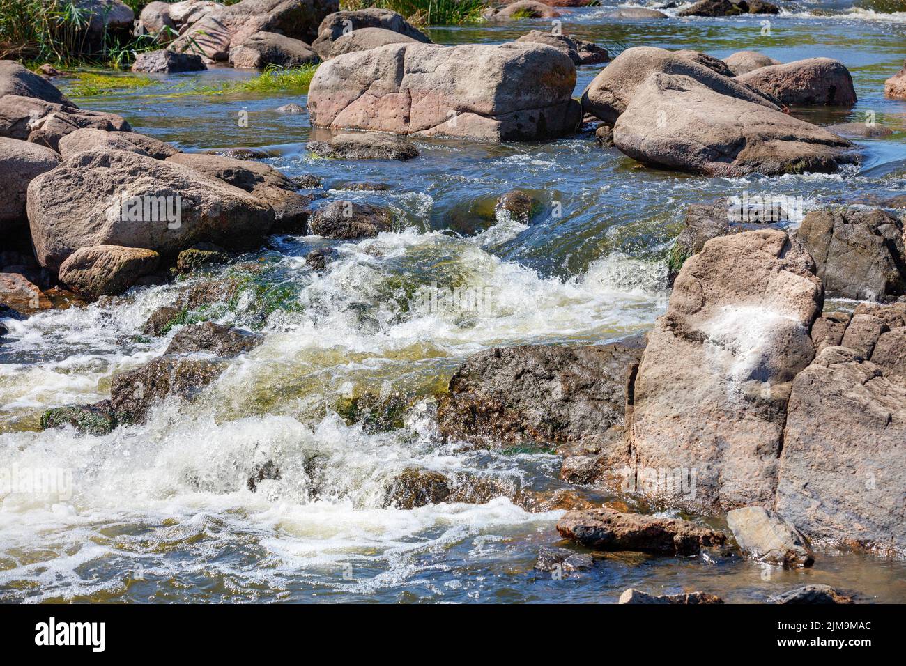 A blue stream of water seethes among rocky stone boulders on a sunny ...