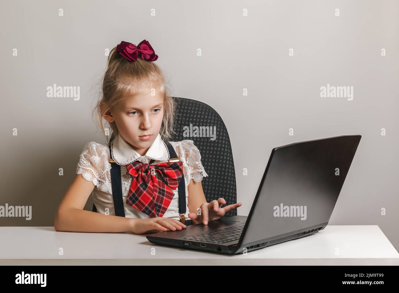 A little girl in a school uniform uses a laptop at a table on a white ...
