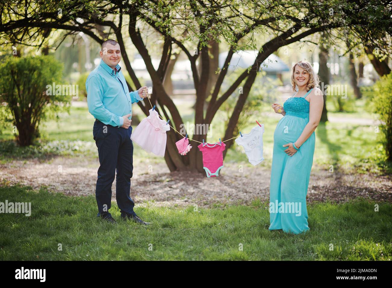 Happy pregnant couple holding girls' baby clothes on string background ...