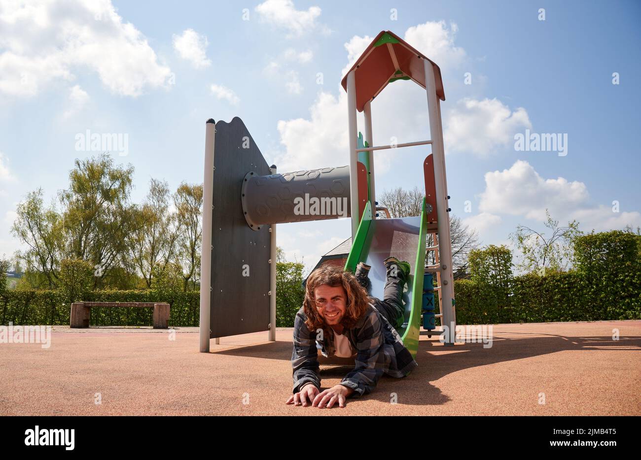 A cheerful young Caucasian man on the kids slides in the playground ...
