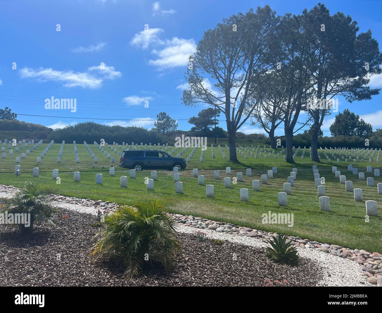 A car is driving along the cemetery road with trees and cloudy sky on ...