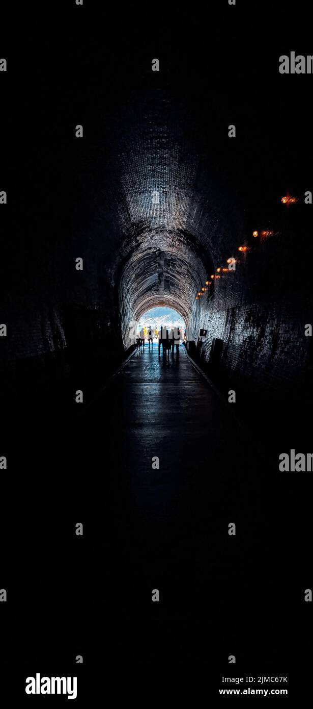 A vertical shot of people walking in a dark narrow tunnel leading to a ...