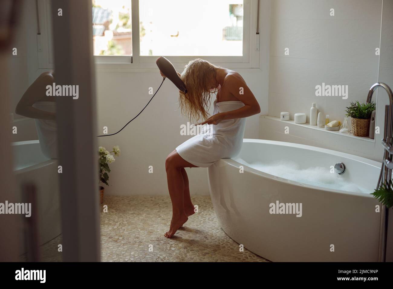 Beautiful Caucasian female drying hair with dryer after taking bath ...