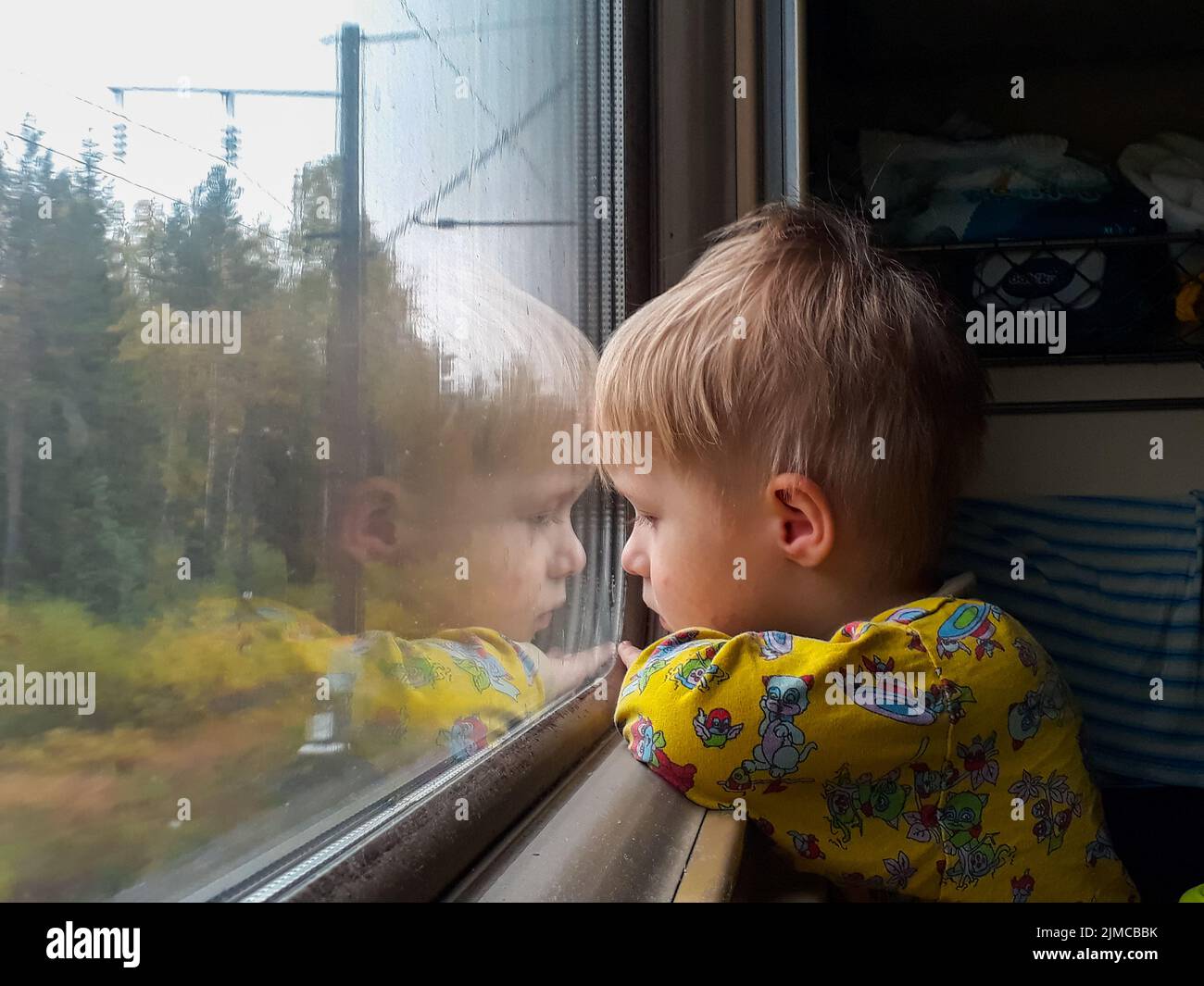 A bright, cute three-year-old boy riding a train looks out the window ...