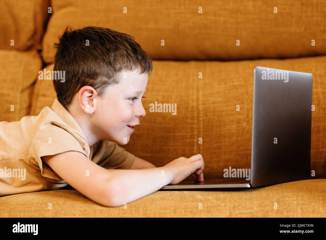 Side view of happy boy lying on a sofa using the laptop while at home ...
