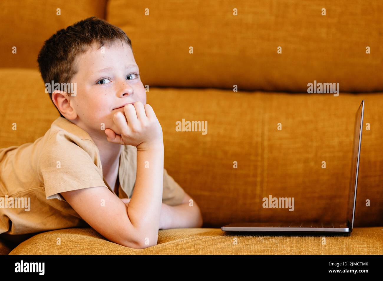 Side view of thoughtful boy lying on a sofa using the laptop while at ...