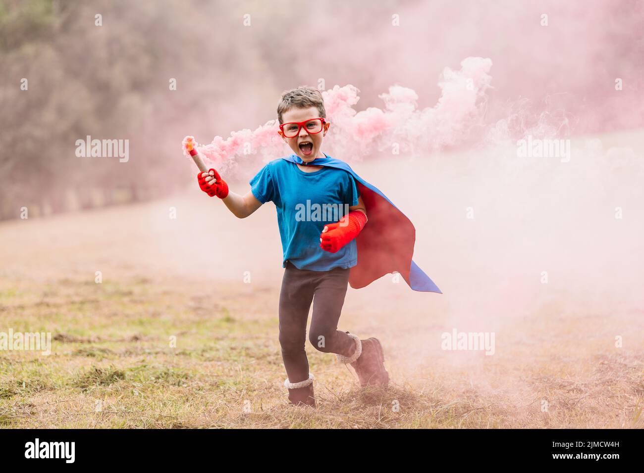 Cute excited boy in superhero costume and glasses holding red smoke ...