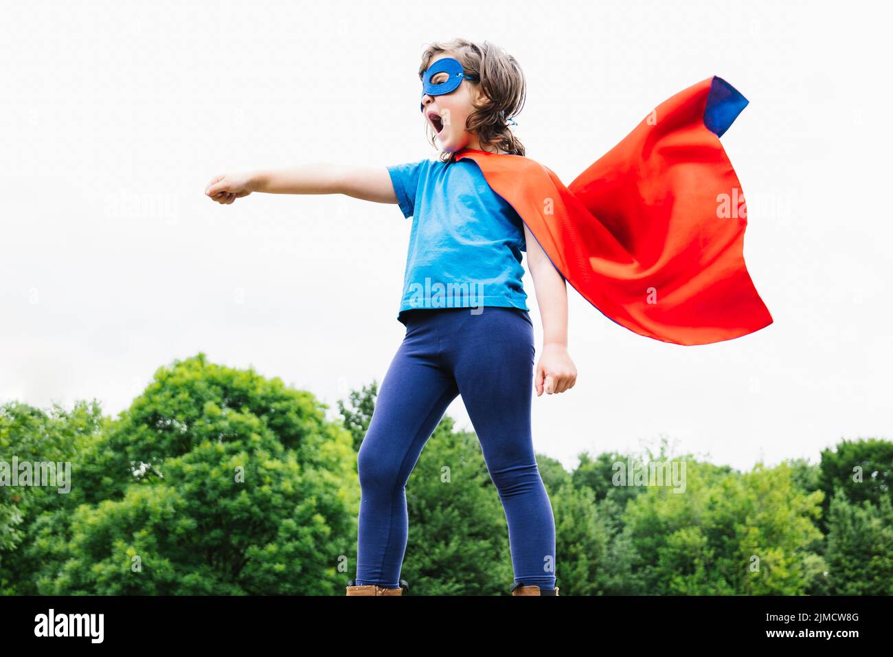 Side view of little female superhero with cape and mask raising arm ...