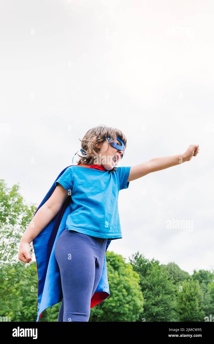 Side view of little female superhero with cape and mask raising arm ...
