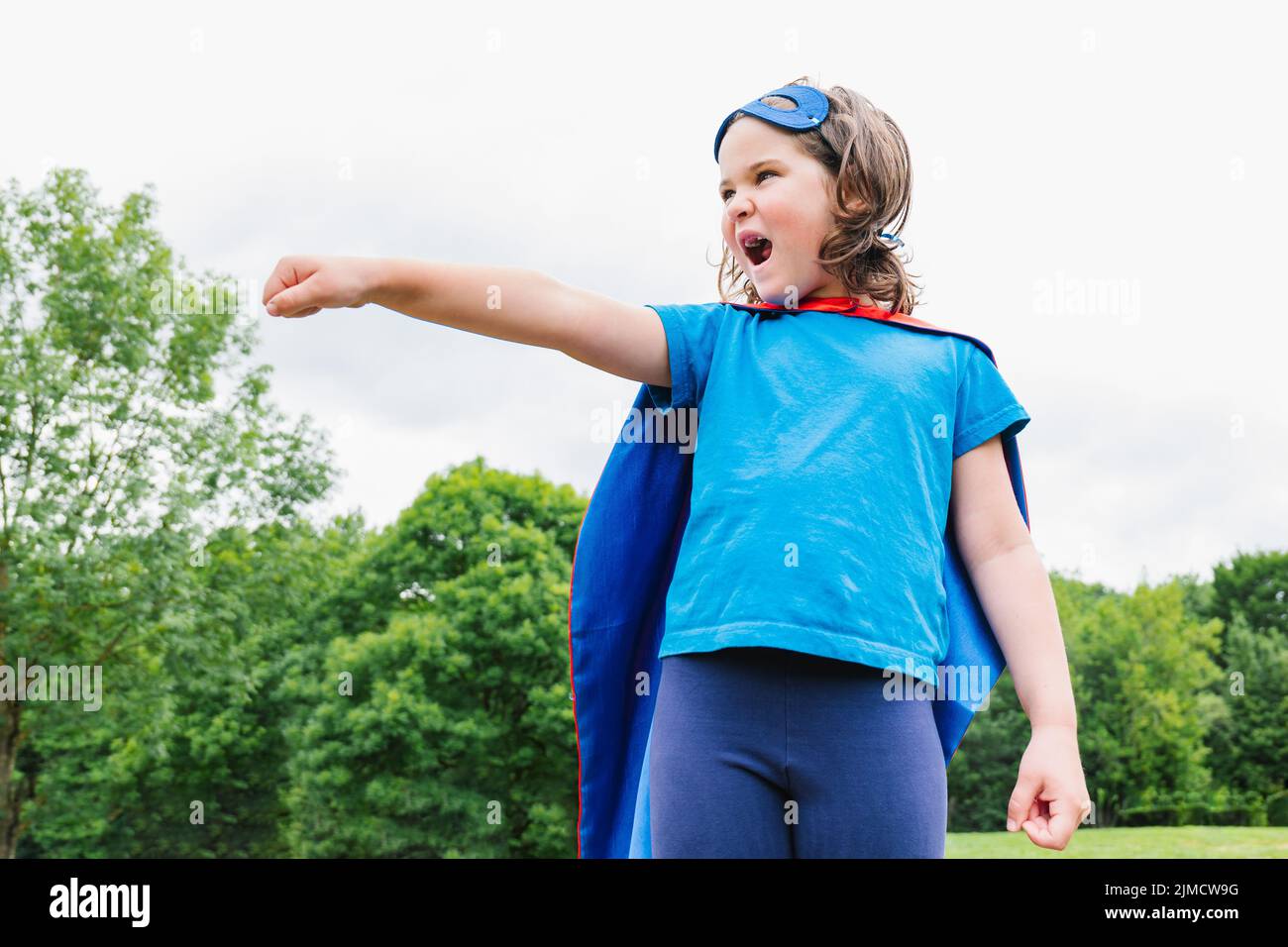 Side view of little female superhero with cape and mask raising arm ...