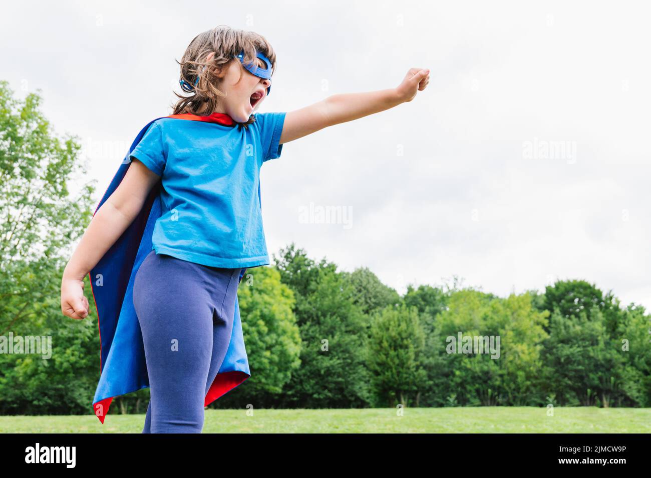 Side view of little female superhero with cape and mask raising arm ...