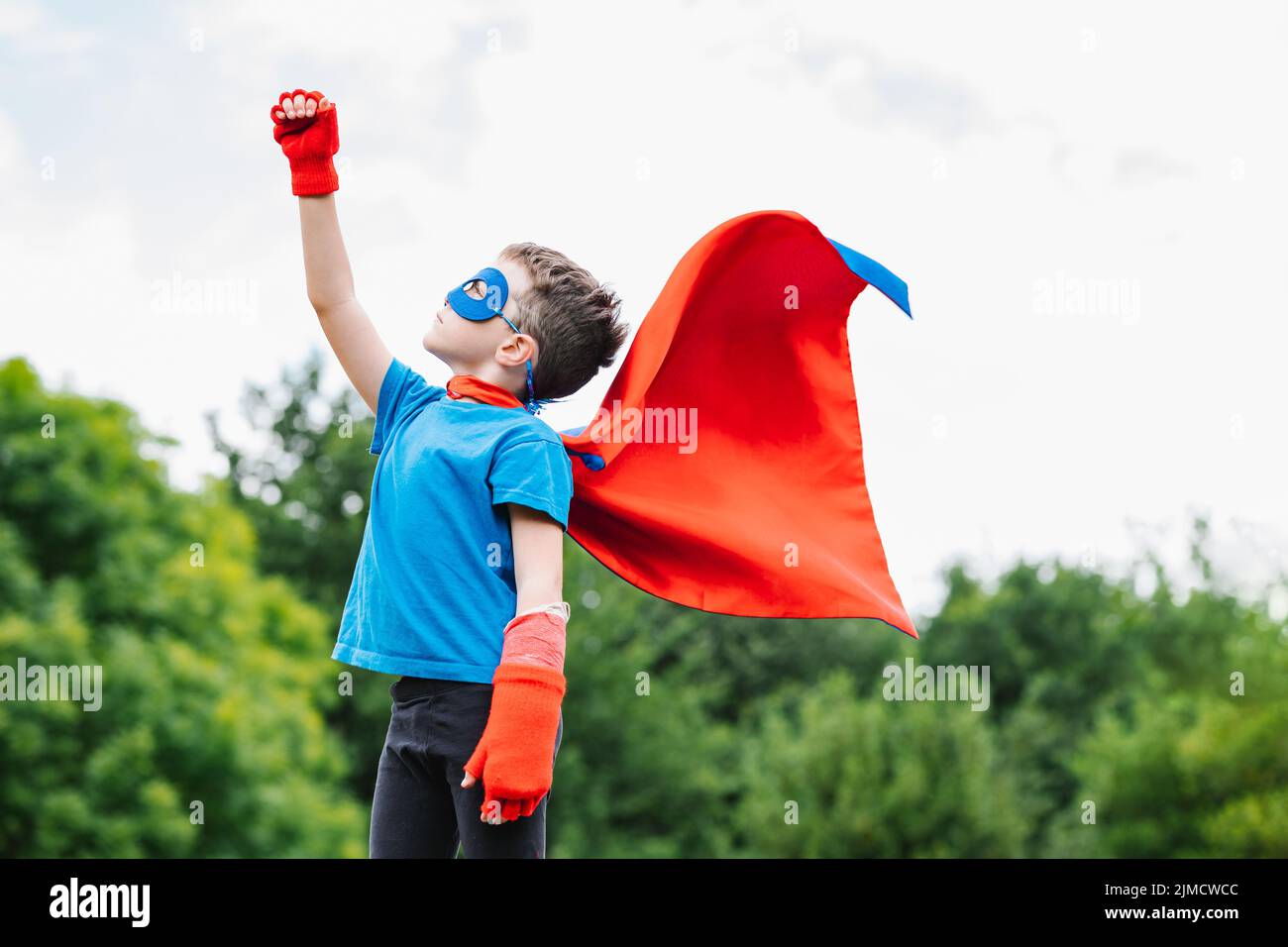 Side view of little superhero with cape and mask raising arm with ...