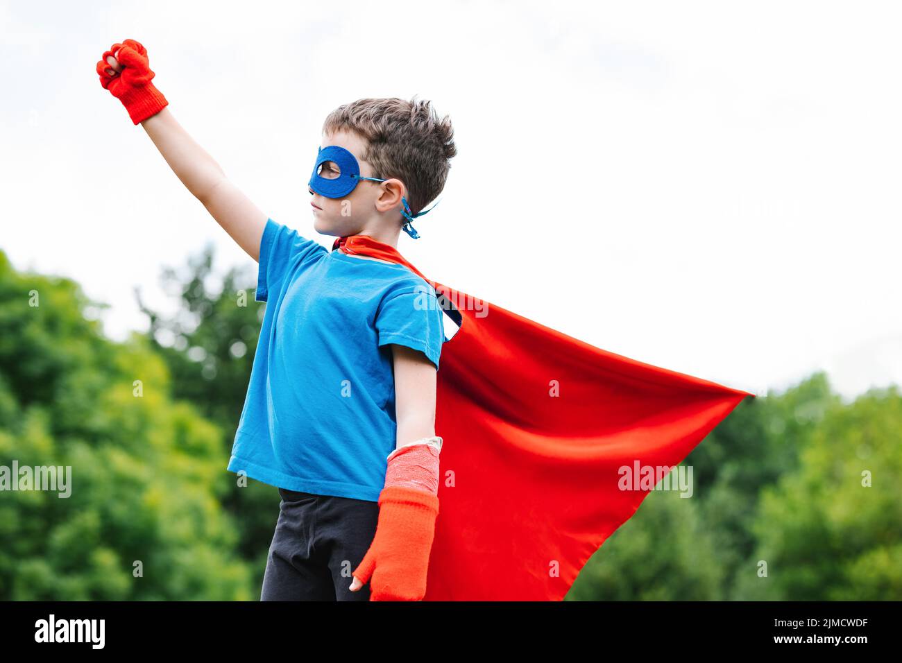 Side view of little superhero with cape and mask raising arm with ...