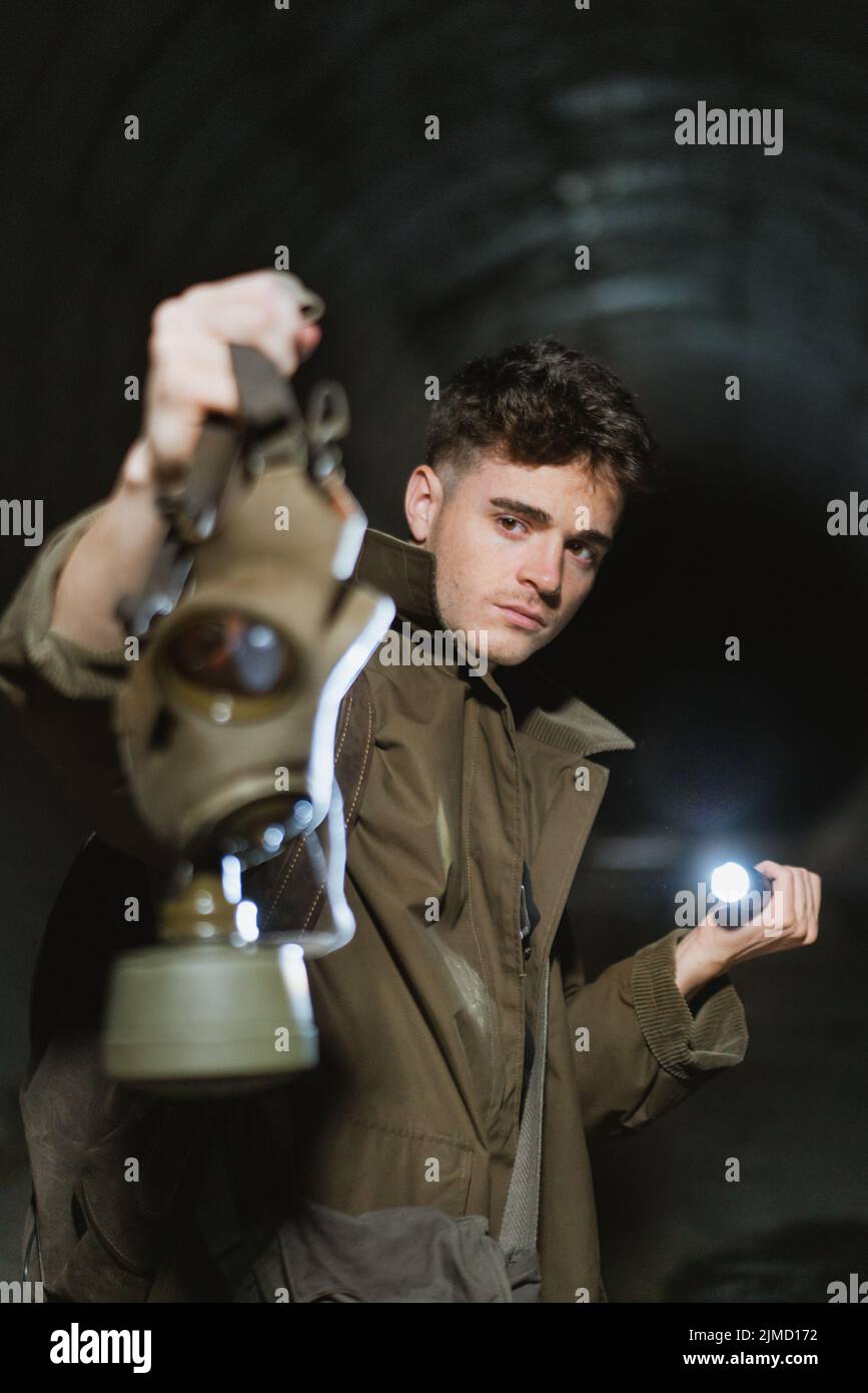Young man in coat illuminating gas mask with flashlight while exploring ...