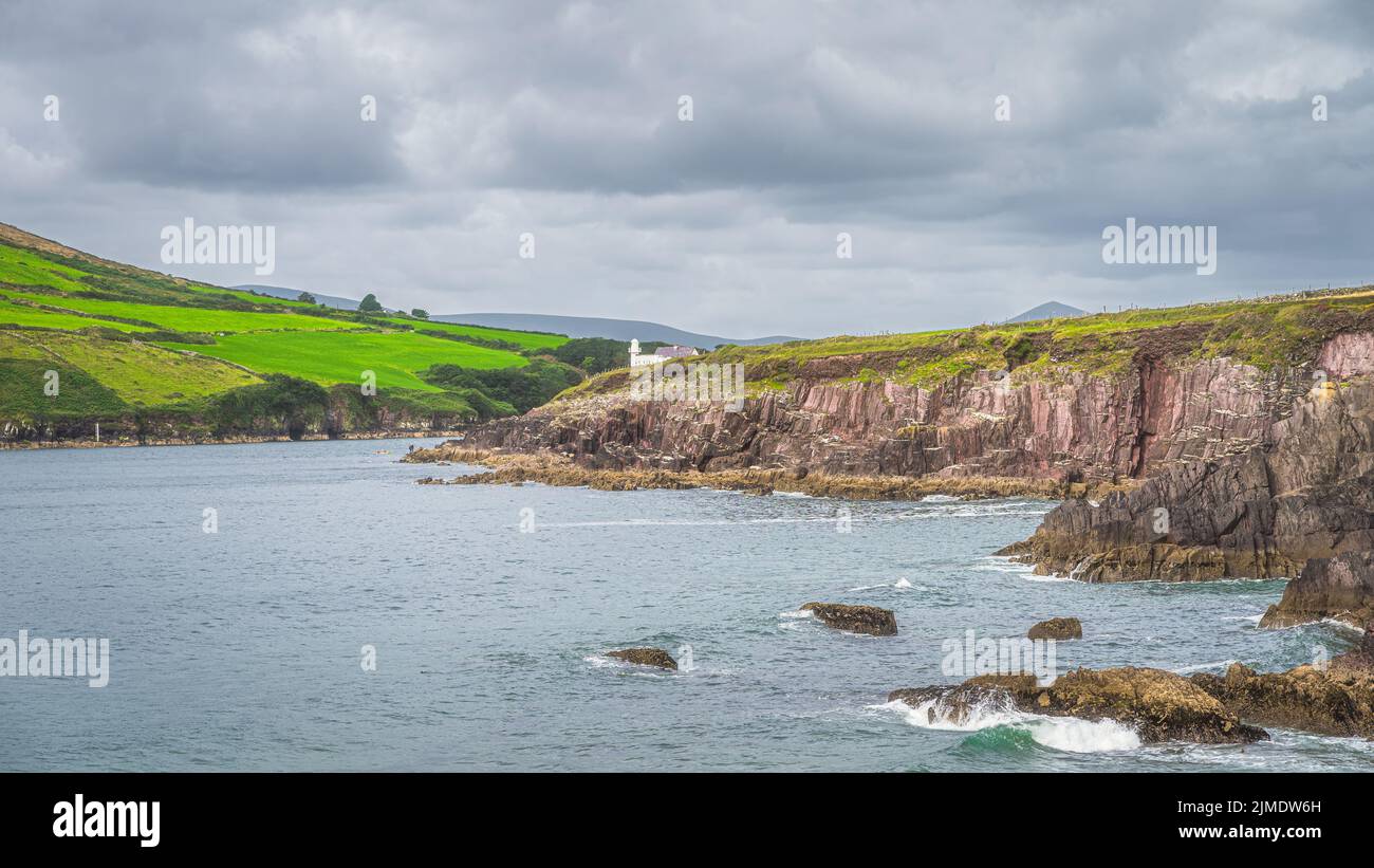 Dingle Lighthouse on the edge of the cliff with green fields in a ...