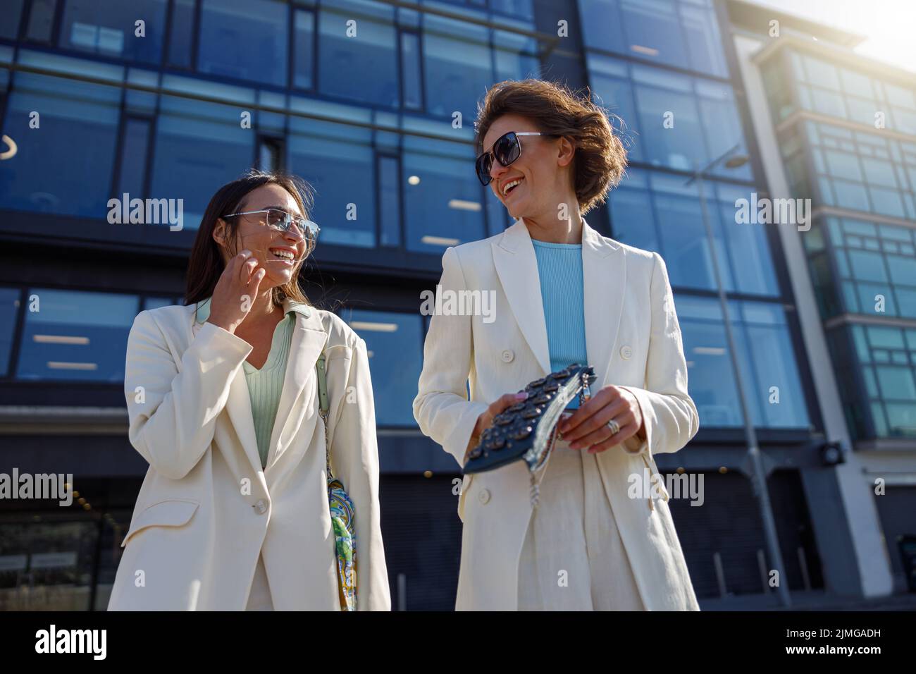 Two smiling woman in trendy outfits laugh and walk outside on ...