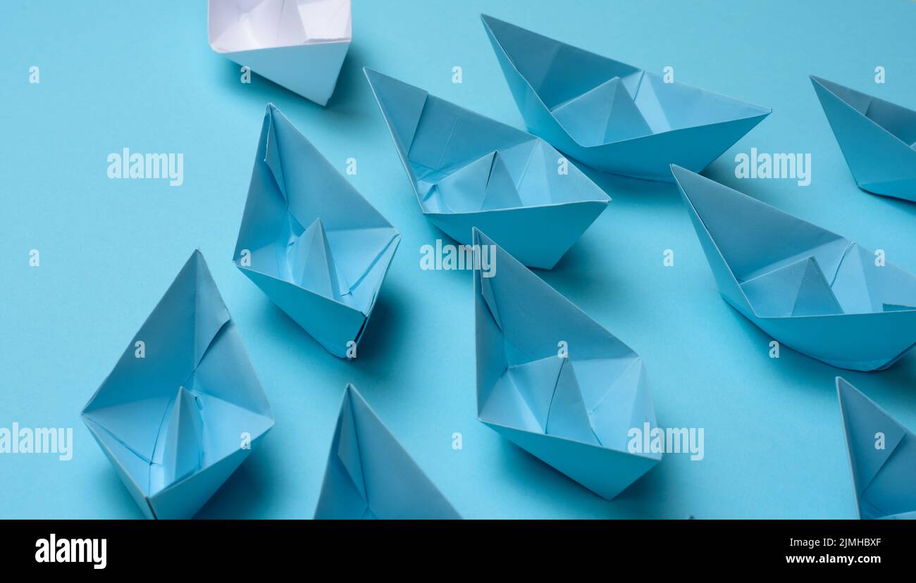 Group of blue paper boats follow white against a light blue background ...