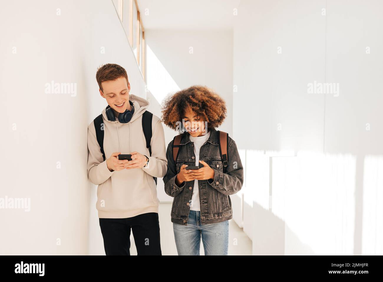 Laughing classmates walking together in corridor holds smartphones ...